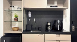 Cozinha compacta com cafeteira, chaleira e pia. Há armários com puxadores e uma pequena cesta sobre o balcão.
