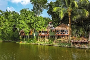 Acomodação às margens do rio, com cabanas de telhado de palha, cercada por vegetação exuberante e um rio tranquilo.