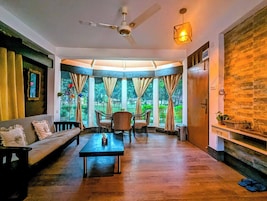 Quarto com sofá, mesa, cadeiras, ventilador de teto e grandes janelas com cortinas.