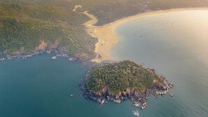 Vista aérea de uma área costeira com uma praia de areia, uma pequena ilha rochosa e um mar azul cristalino.
