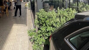 Uma área de estar coberta ao ar livre, com mesas e cadeiras, um carro estacionado e um edifício com sacadas ao fundo.