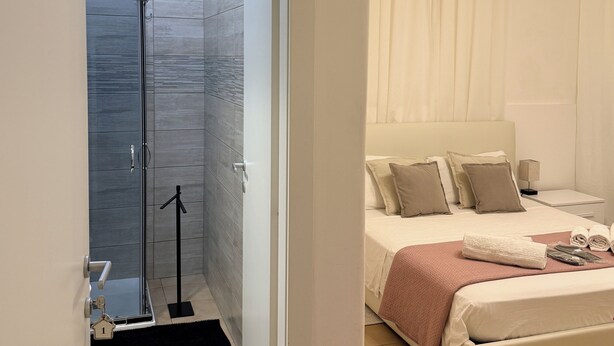 Quarto de hotel moderno com uma cama grande, uma mesinha de cabeceira e um criado-mudo. Possui um banheiro com chuveiro e porta de vidro.