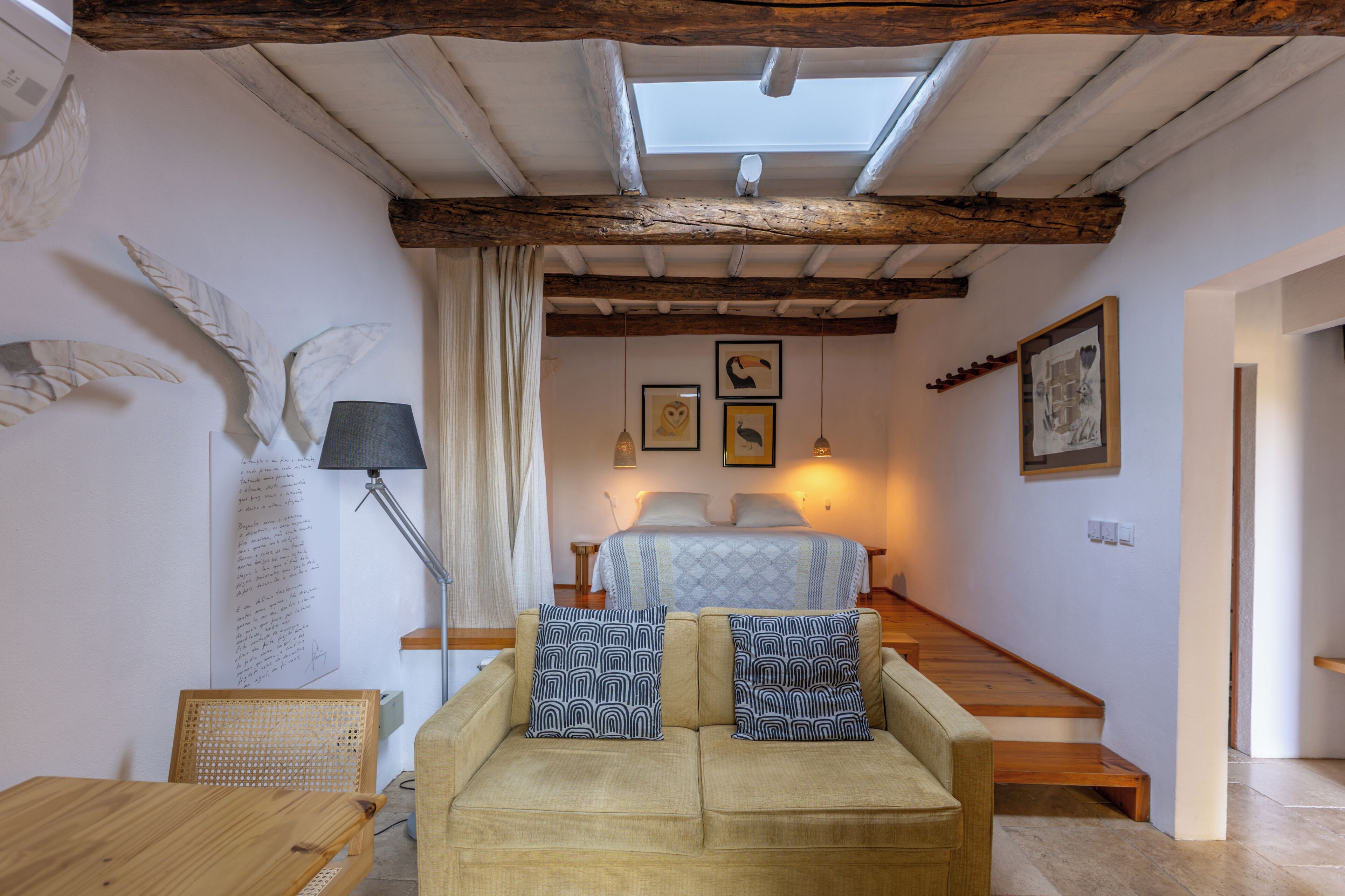 Quarto aconchegante com cama, sofá e escrivaninha. Há um claraboia acima da cama e uma luminária sobre a escrivaninha.
