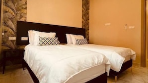 Cama de casal com estrutura preta e roupa de cama branca, uma pequena mesa de cabeceira e parede com papel de parede estampado com folhas.