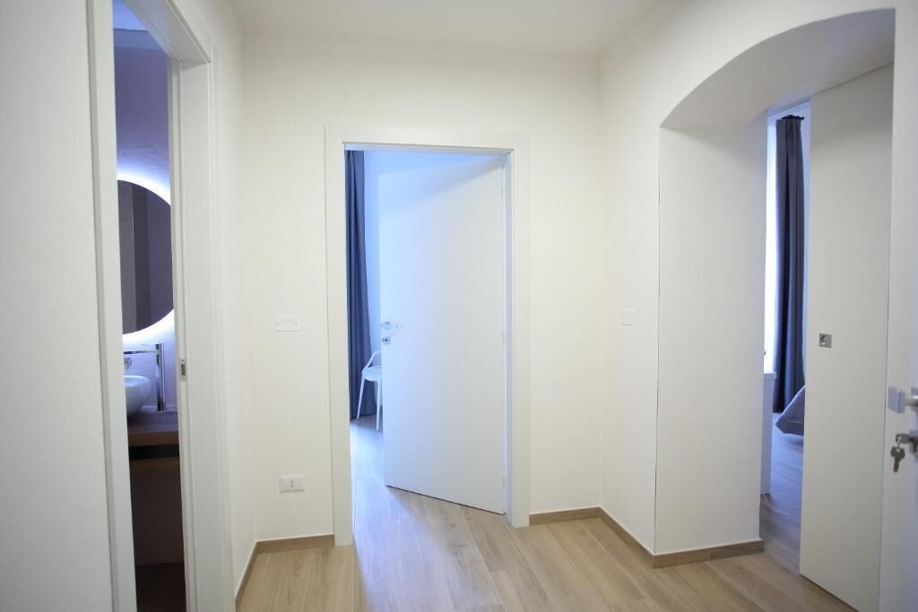 Um corredor com paredes brancas e soalho de madeira, que leva a um quarto com cama e janela.