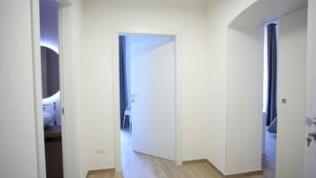Um corredor com paredes brancas e soalho de madeira, que leva a um quarto com cama e janela.