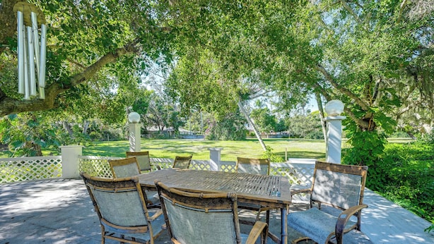 Um pátio externo com mesa e cadeiras, rodeado por vegetação exuberante e vista para um gramado e árvores.