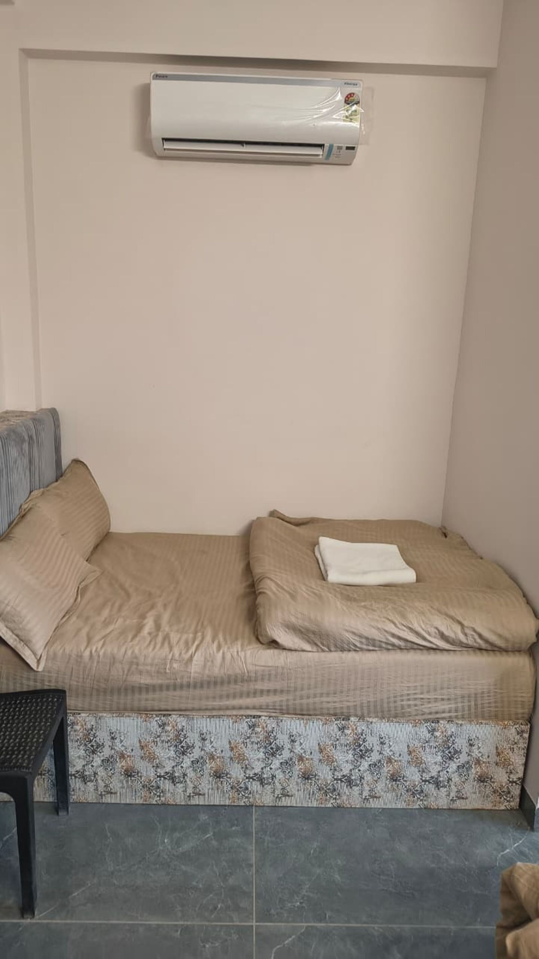 Quarto pequeno com uma cama de solteiro, colchão bege, travesseiro branco e tapete estampado.