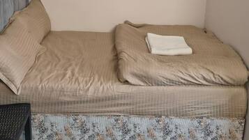 Quarto pequeno com uma cama de solteiro, colchão bege, travesseiro branco e tapete estampado.