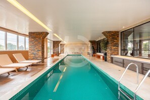 Piscina interior com espreguiçadeiras, detalhes em pedra e grandes janelas.