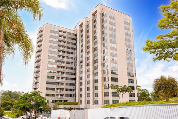 Um edifício residencial com várias andares e varandas, cercado por vegetação e um céu claro.
