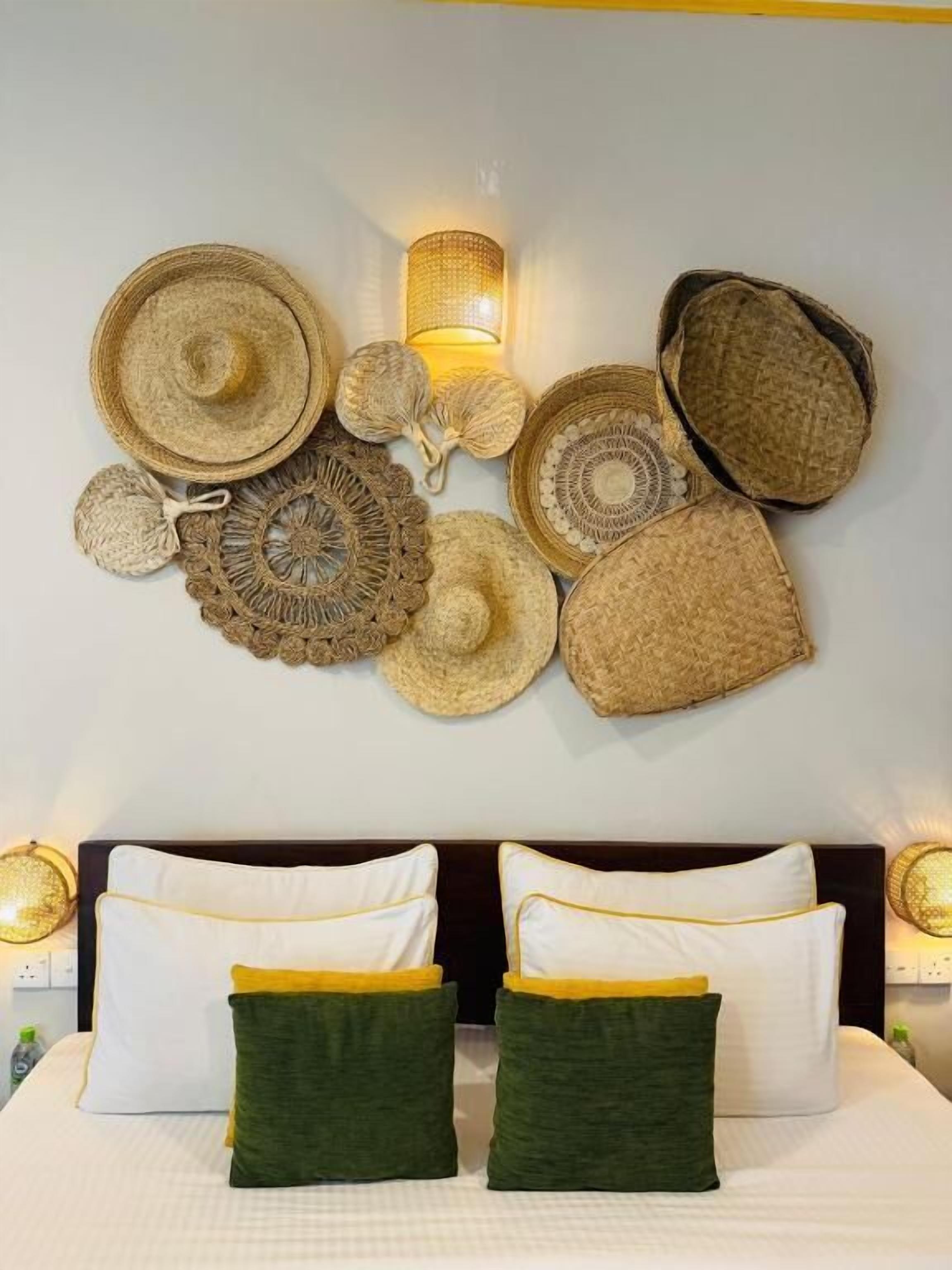Cama com almofadas brancas e verdes, cabeceira escura e parede decorada com cestos de tecido e uma luminária de parede.