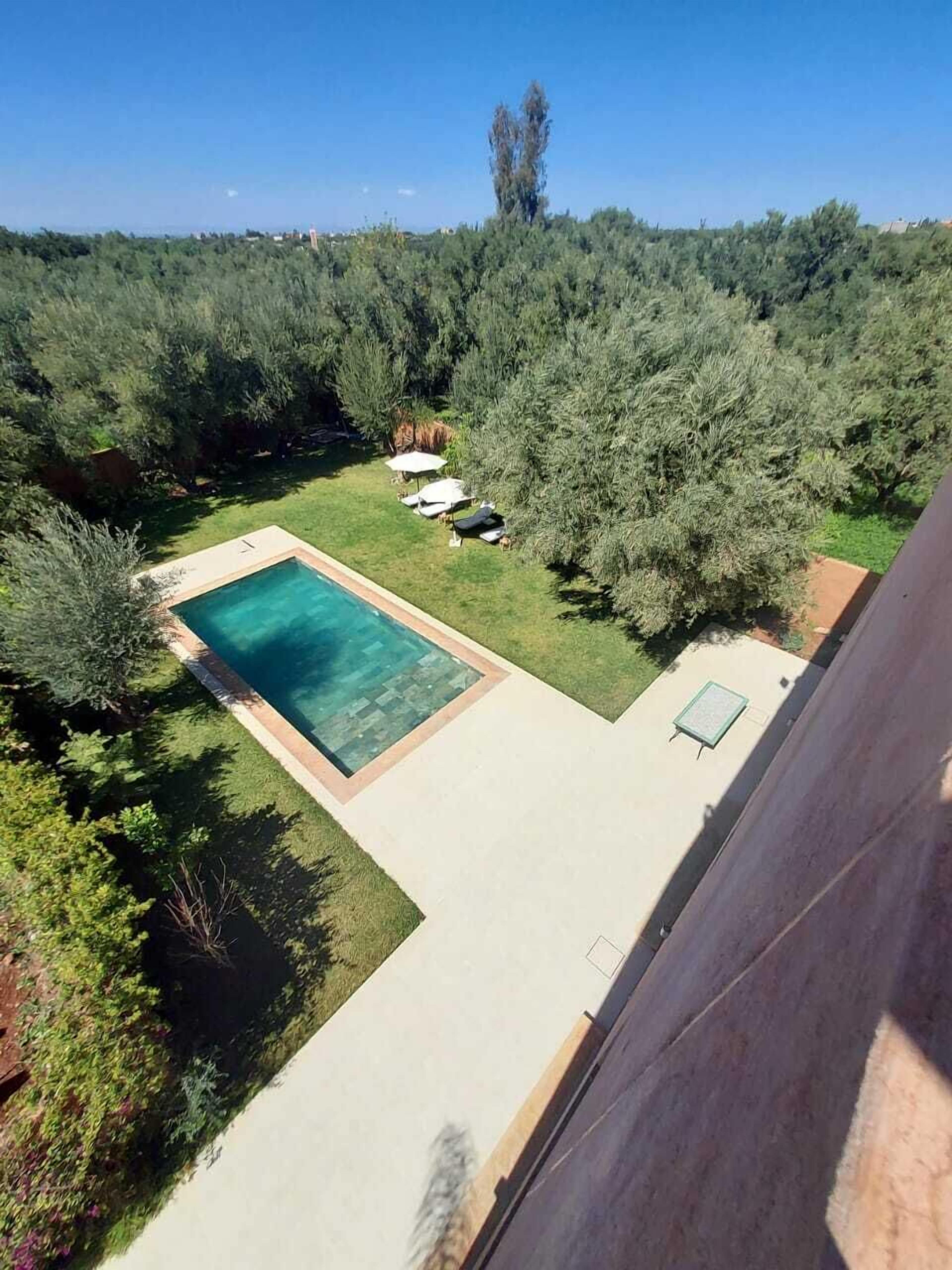Uma piscina rodeada por um espaço gramado com árvores, um alpendre com mesa e cadeiras, e vista para a paisagem circundante.