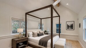 Uma cama com dossel e colunas, acompanhada de uma mesinha de cabeceira e uma mesa lateral, com vista para o exterior através de amplas janelas.
