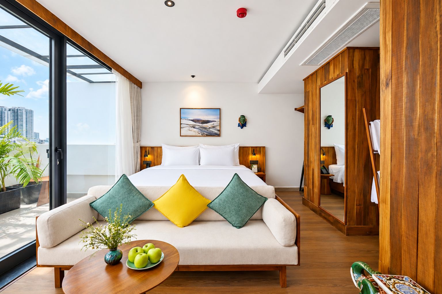 Quarto de hotel moderno com uma cama grande, um sofá, uma pequena mesa com maçãs e vista para a cidade.