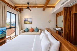 Quarto de hotel com uma cama grande, um banco de madeira, uma televisão, um quadro e vista para o exterior.