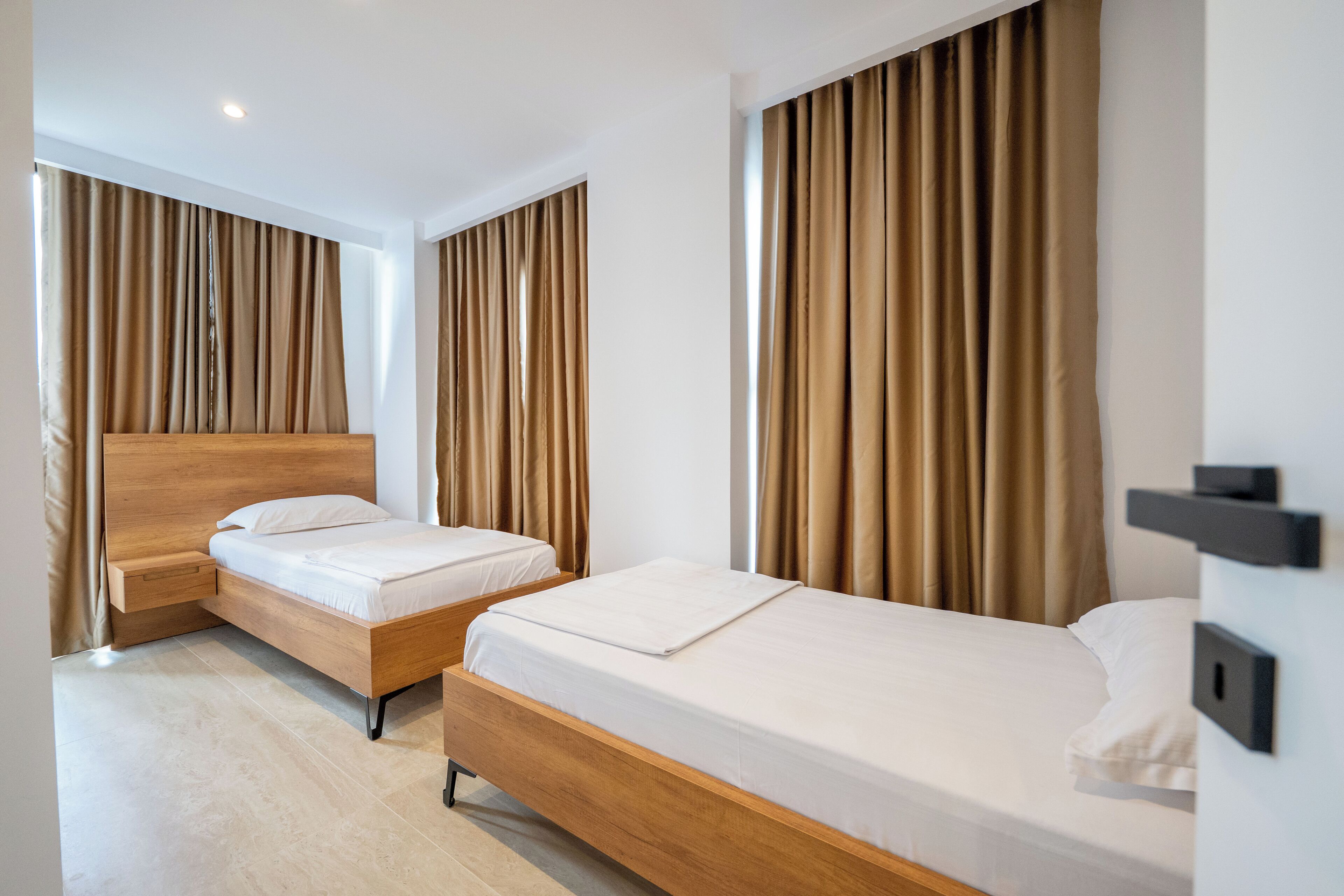 Quarto de hotel com duas camas, cabeceiras de madeira, cortinas bege e parede branca com interruptor de luz.