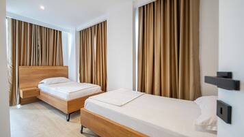 Quarto de hotel com duas camas, cabeceiras de madeira, cortinas bege e parede branca com interruptor de luz.