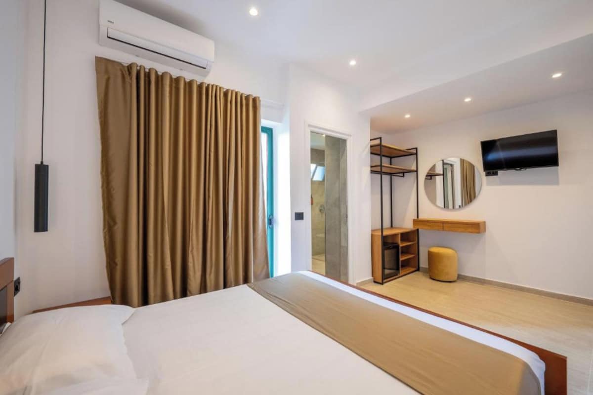 Quarto de hotel moderno com uma cama grande, televisão na parede e um pequeno espaço de estar com um espelho.