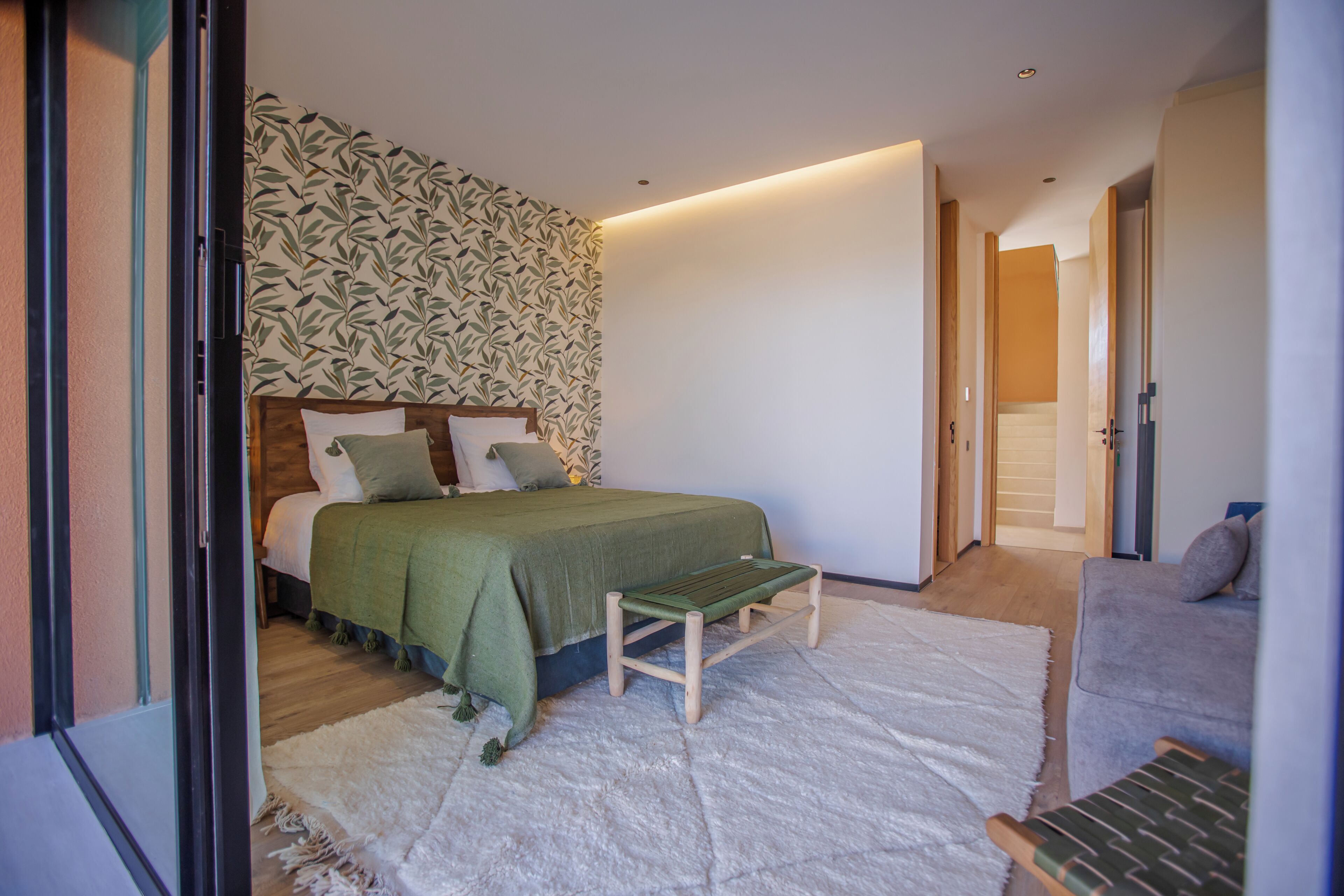 Quarto moderno com cama, um banquinho, um sofá e parede estampada.