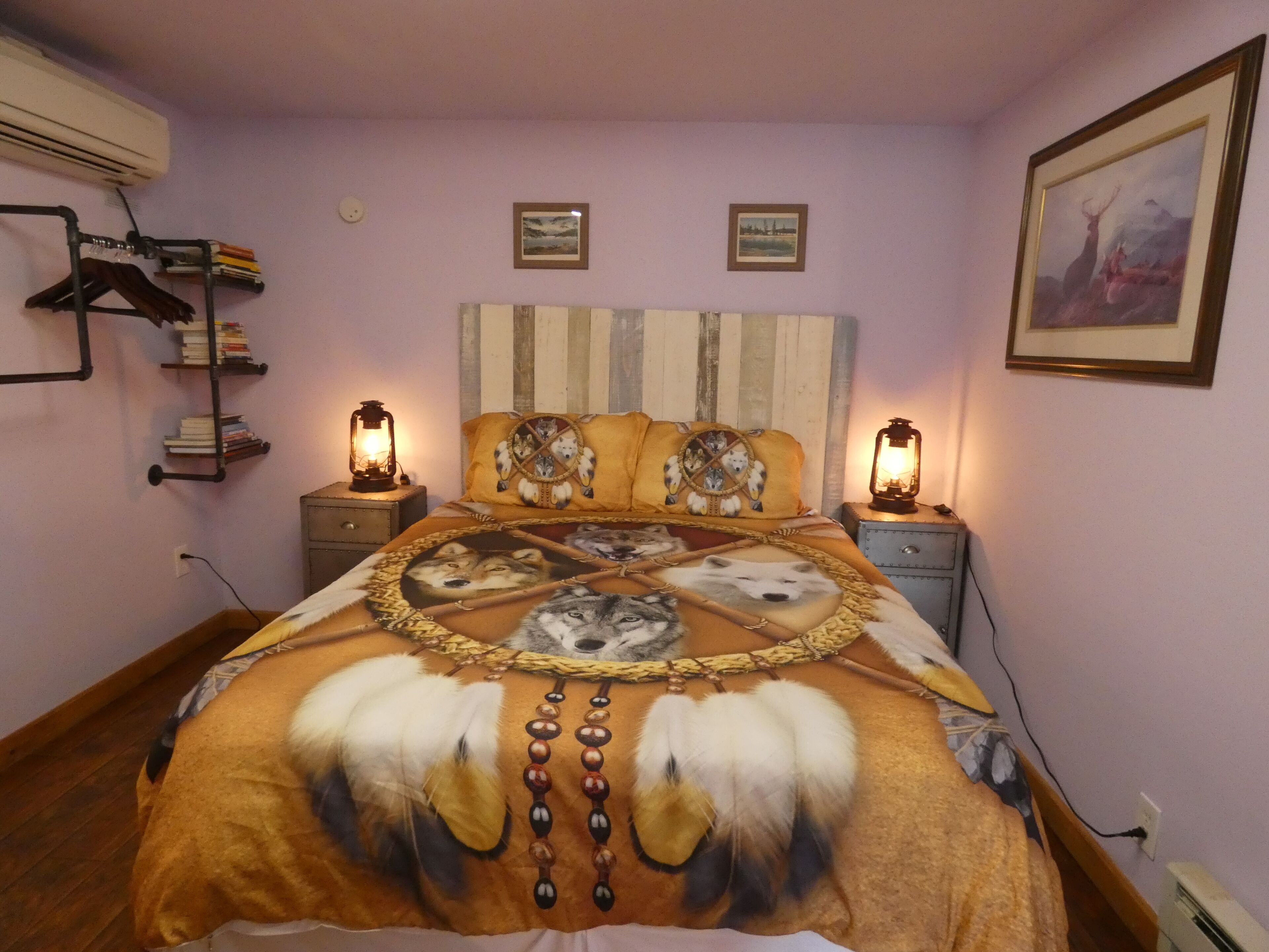 Quarto com cama, edredom com tema de lobo, duas luminárias de apoio, prateleira na parede com livros e um quadro emoldurado sobre a cama.