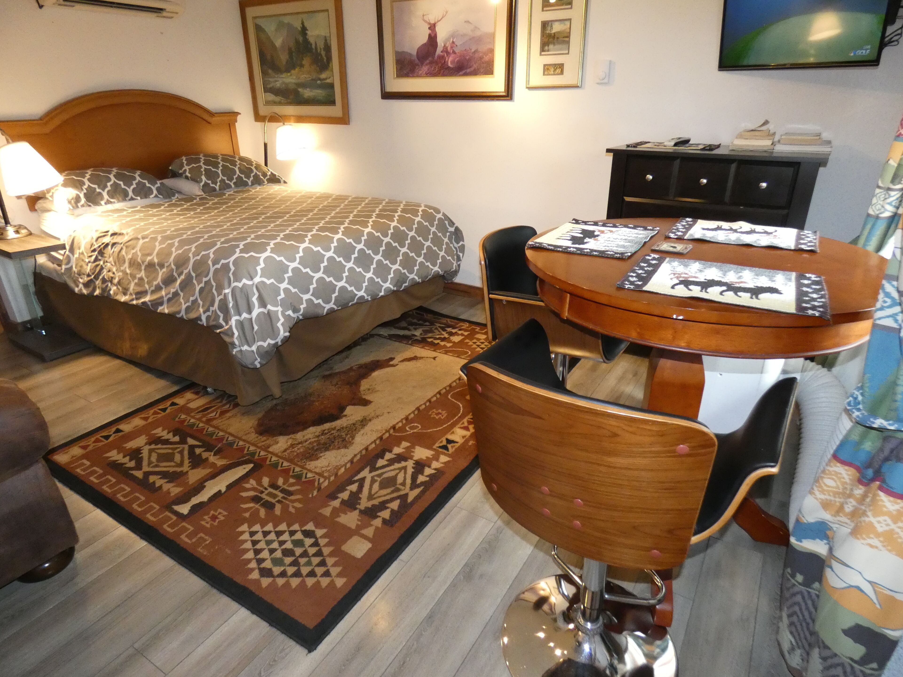 Quarto com cama, escrivaninha e cadeira. Há um tapete no chão e uma televisão na parede.