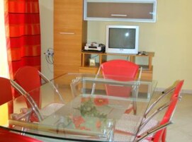 Uma sala de estar moderna com uma mesa de centro de vidro, cadeiras vermelhas, uma televisão de tela plana e um armário de madeira.