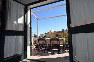 Uma porta aberta que dá para um pátio externo com uma mesa e cadeiras, um guarda-sol amarelo e vista para um edifício ao fundo.