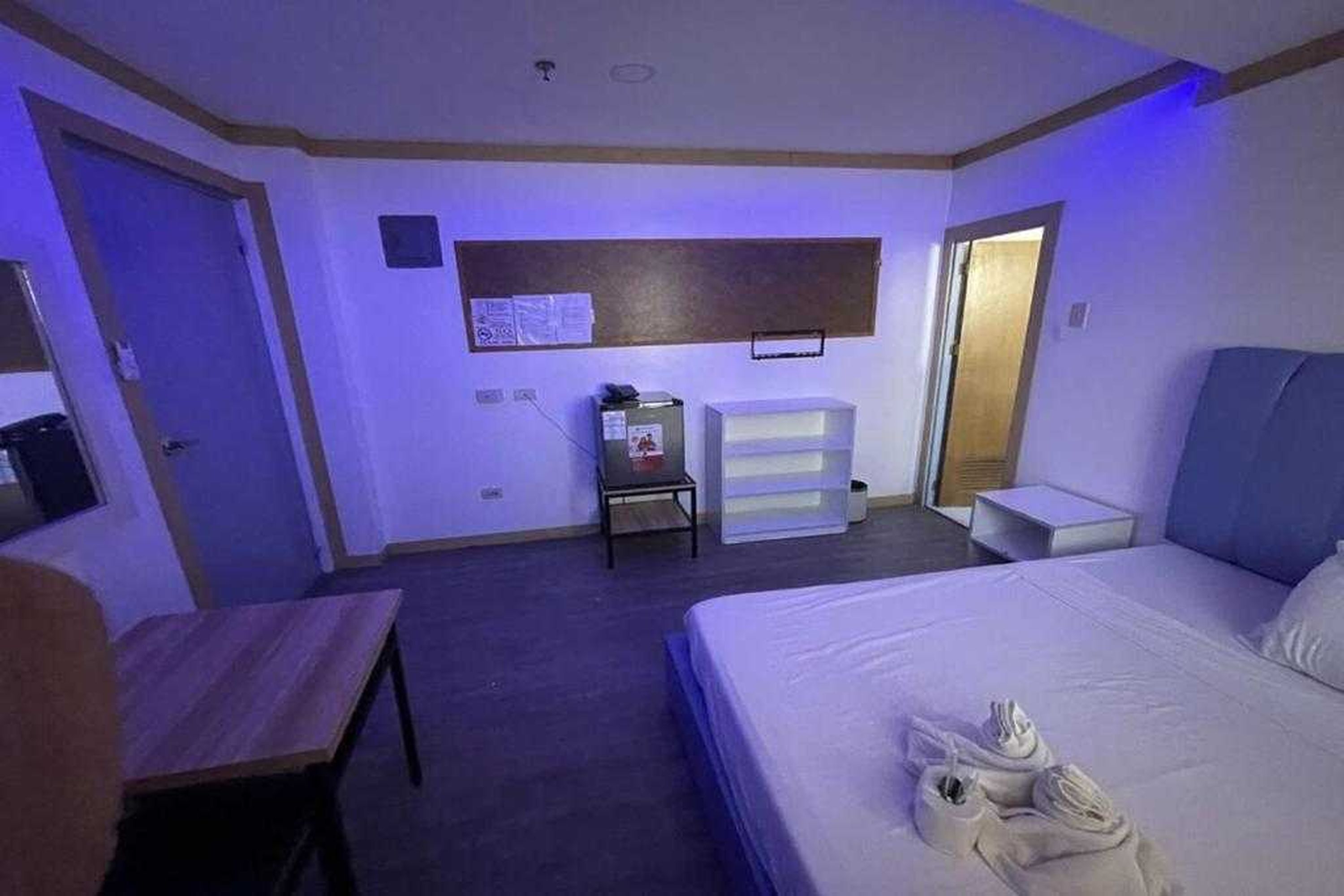 Quarto de hotel com cama, mesa de cabeceira e escrivaninha. Há uma cadeira, uma mesinha e um armário.