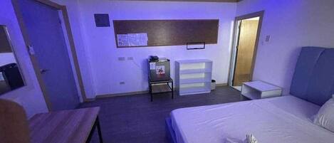 Quarto de hotel com cama, mesa de cabeceira e escrivaninha. Há uma cadeira, uma mesinha e um armário.