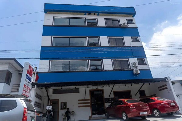 Um edifício de vários andares com fachada azul e branca, um letreiro com 'JM Lodge' e dois carros vermelhos estacionados em frente.