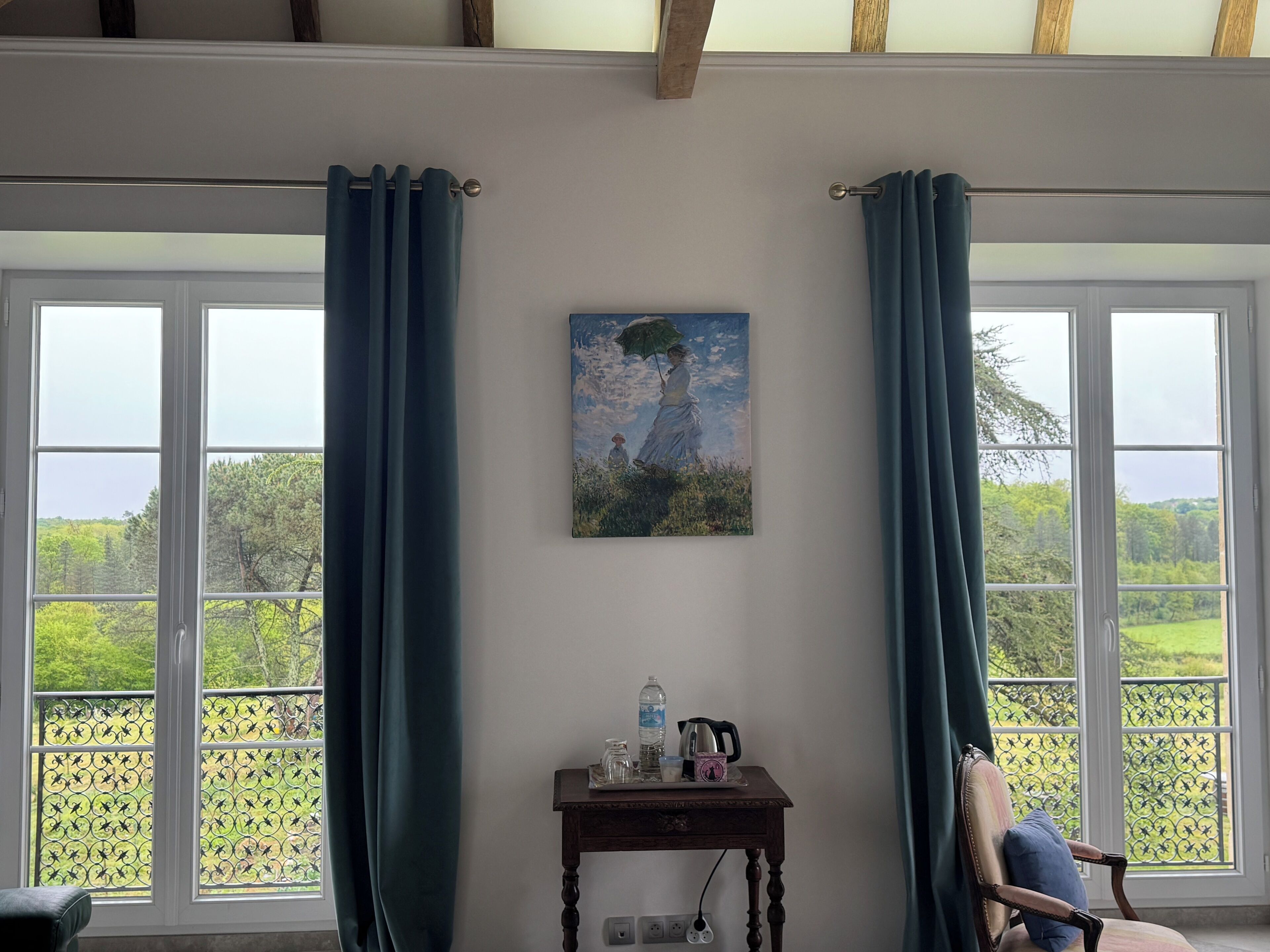 Quarto com uma janela grande, um quadro na parede, uma mesa com uma chaleira e uma garrafa, e uma cadeira.