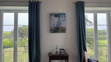 Quarto com uma janela grande, um quadro na parede, uma mesa com uma chaleira e uma garrafa, e uma cadeira.