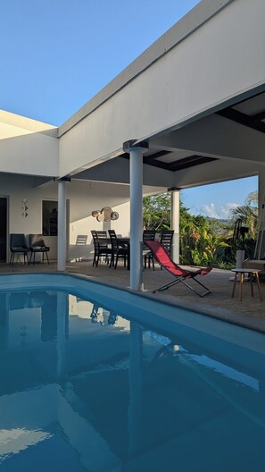 Uma área moderna de piscina ao ar livre, com um espaço de estar coberto, uma espreguiçadeira vermelha e vista para o verde.