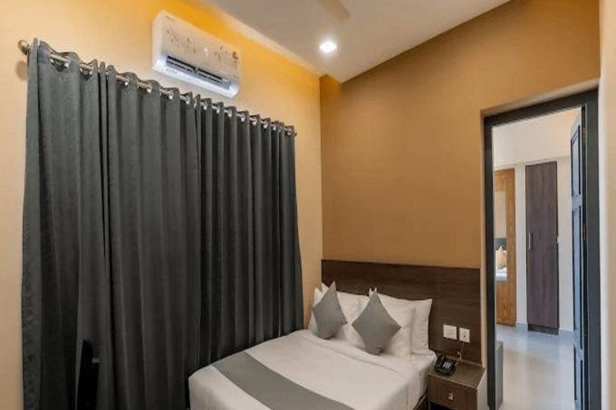 Quarto de hotel com cama, cortinas, ventilador de teto, ar condicionado, mesa de cabeceira com telefone e porta que dá para outro cômodo.
