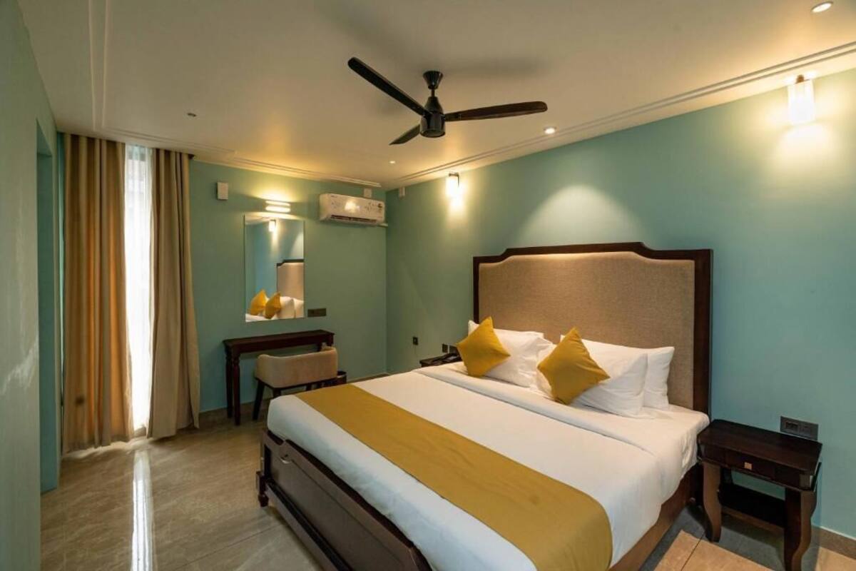 Quarto de hotel com cama, ventoinha no teto, mesa de cabeceira, escrivaninha, cadeira, espelho e janela com cortinas.