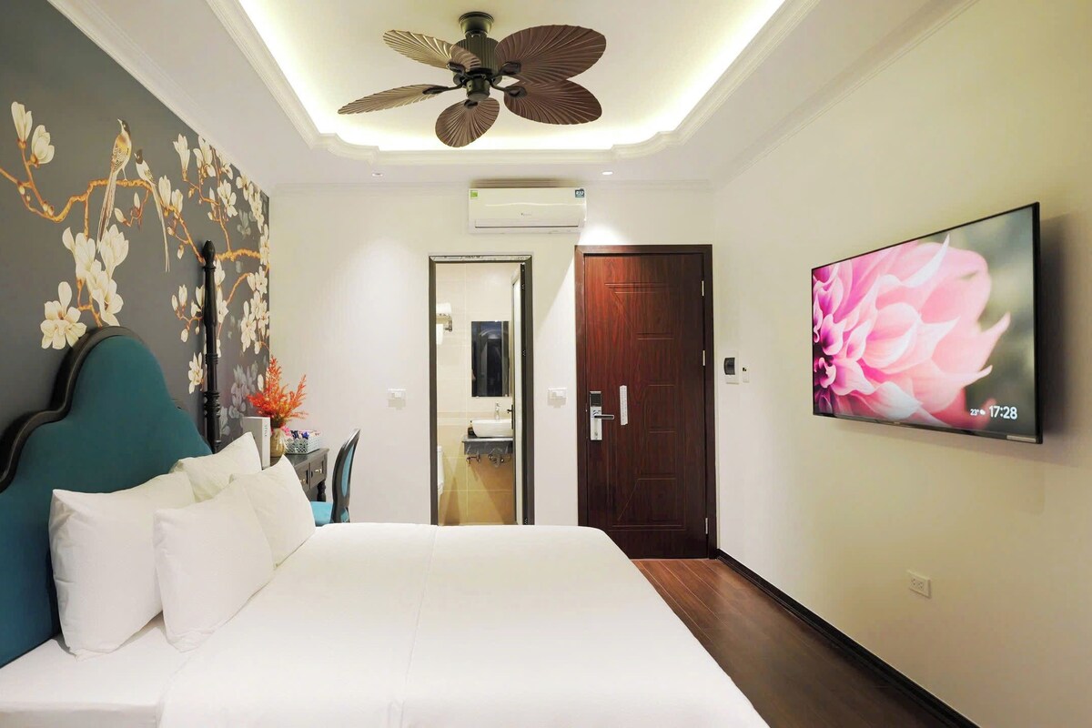 Quarto de hotel com uma cama, um ventilador de teto, uma televisão na parede e uma porta que dá para um banheiro.