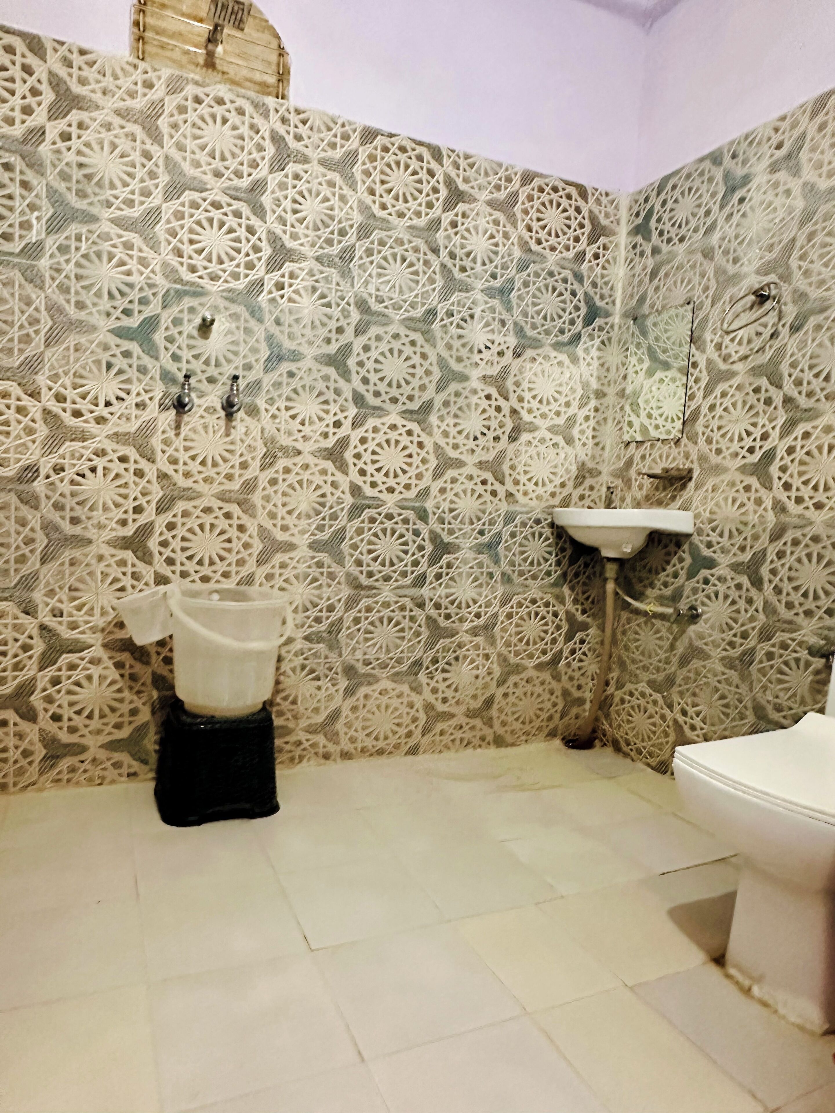 Casa de banho com parede de azulejos estampados, sanita, lavatório e um contentor de lixo preto.