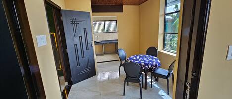 Um quarto com soalho em azulejos, uma mesa com cadeiras, uma porta com design geométrico e uma janela com vista para o verde.
