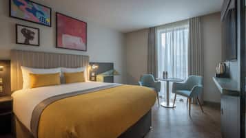 Quarto de hotel com uma cama grande, duas cadeiras, uma mesinha e uma televisão.