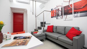 Uma sala de estar moderna com um sofá cinzento, almofadas vermelhas, uma mesa branca com revistas, uma porta vermelha e arte abstrata na parede.