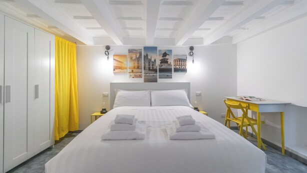 Cama bem arrumada com lençóis e toalhas brancas, uma escrivaninha com cadeira e uma cortina amarelo brilhante.