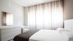 Quarto de hotel moderno com uma cama grande, uma mesa de cabeceira, uma cadeira e um guarda-roupa.