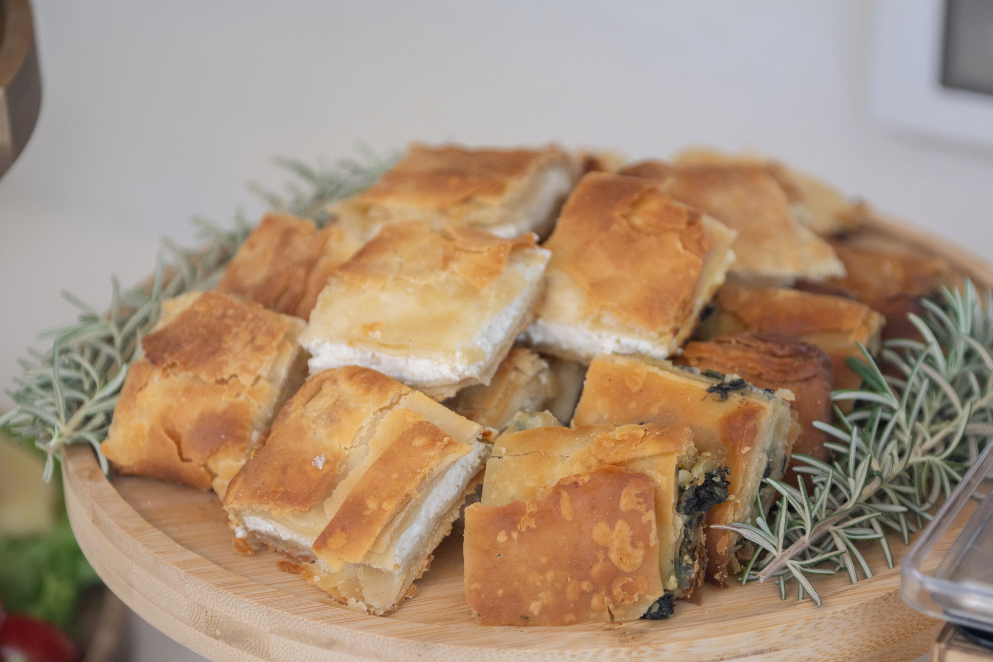 Uma tábua de madeira com vários pedaços de pastelaria assada, possivelmente uma espécie de torta salgada, guarnecida com alecrim.