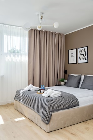 Quarto de hotel moderno com uma cama grande, uma mesinha de cabeceira com uma planta e vista para o exterior através de cortinas leves.
