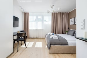 Quarto de hotel moderno com cama, mesa de cabeceira, cadeira, televisão, cortinas e janela.