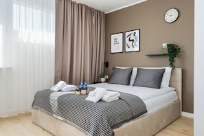 Uma cama bem arrumada com colcha xadrez, uma mesinha de cabeceira com bandeja de amenities e um relógio de parede.