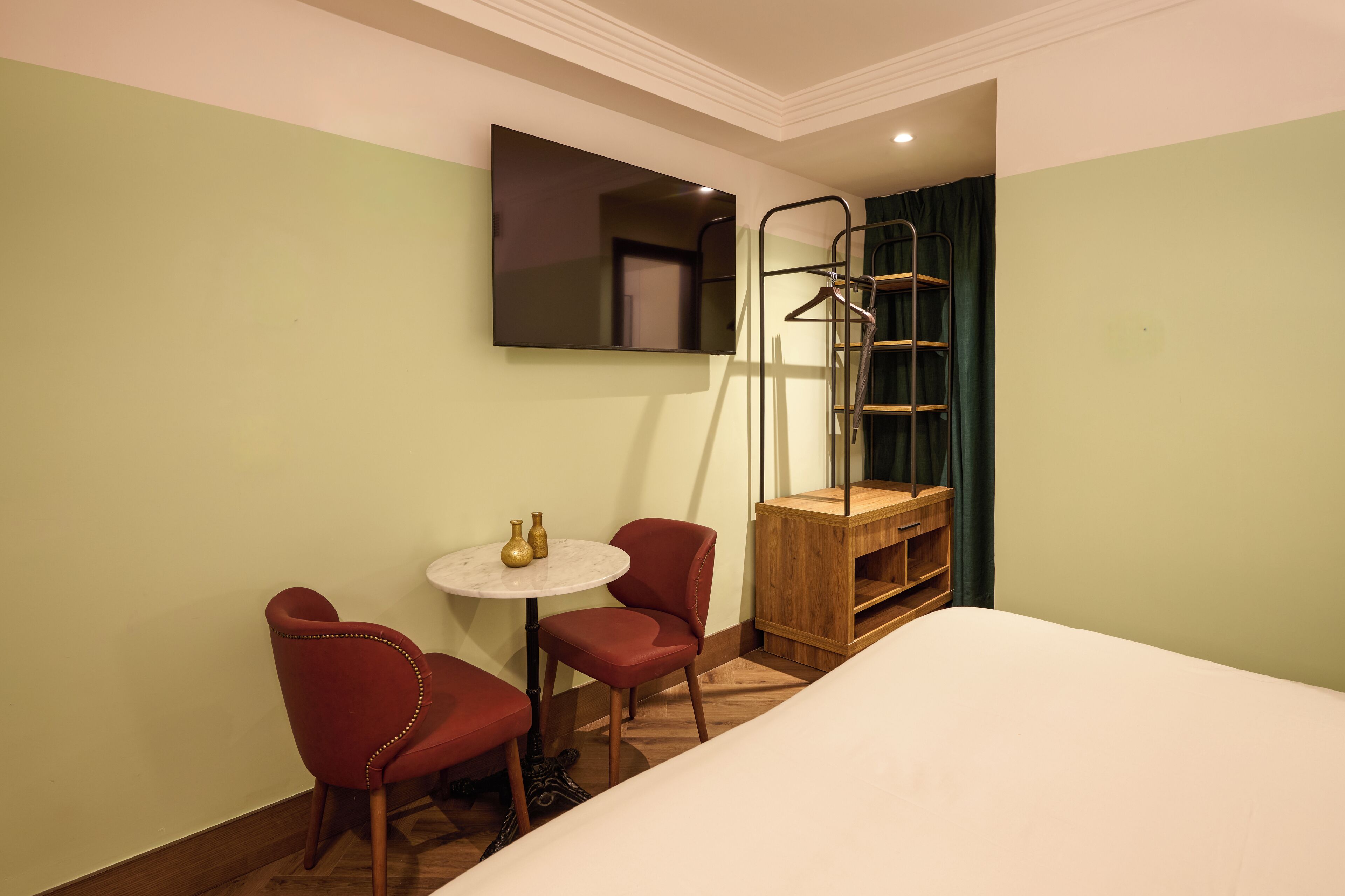 Quarto de hotel com uma cama, duas cadeiras vermelhas, uma mesa redonda pequena com dois vasos, uma estante de madeira e uma televisão fixada na parede.