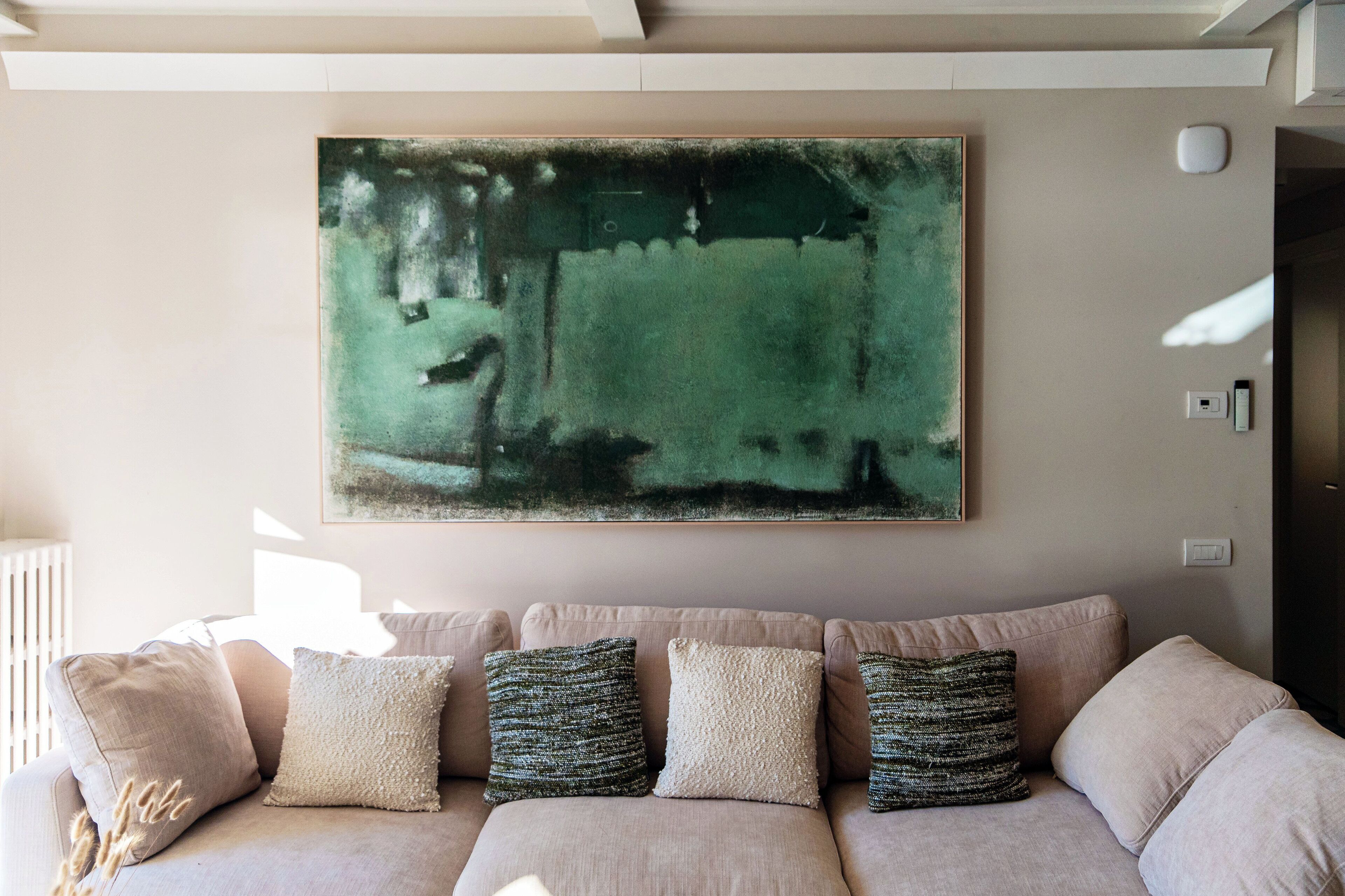 Uma sala de estar com sofá bege, uma grande pintura abstrata e uma janela que ilumina naturalmente o espaço.