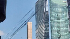 Um edifício moderno de arranha-céu com fachada de vidro, um edifício menor adjacente e uma área arborizada em primeiro plano.